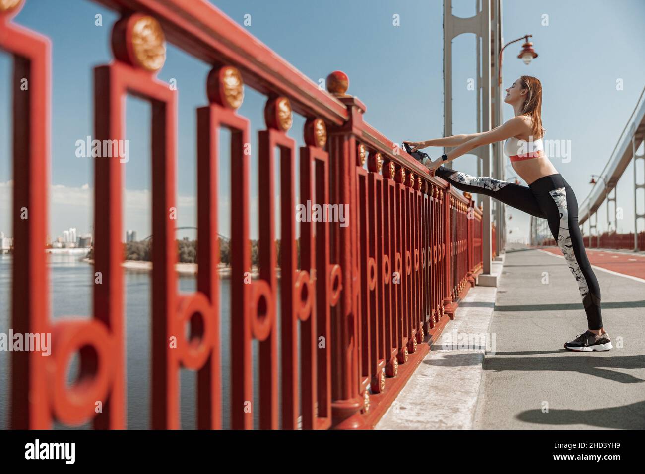 Positive woman does stretching exercise for legs near railing on bridge ...