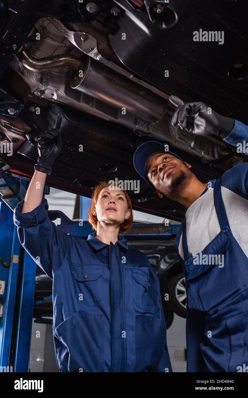 Low angle view of multiethnic mechanics pointing at car bottom in ...