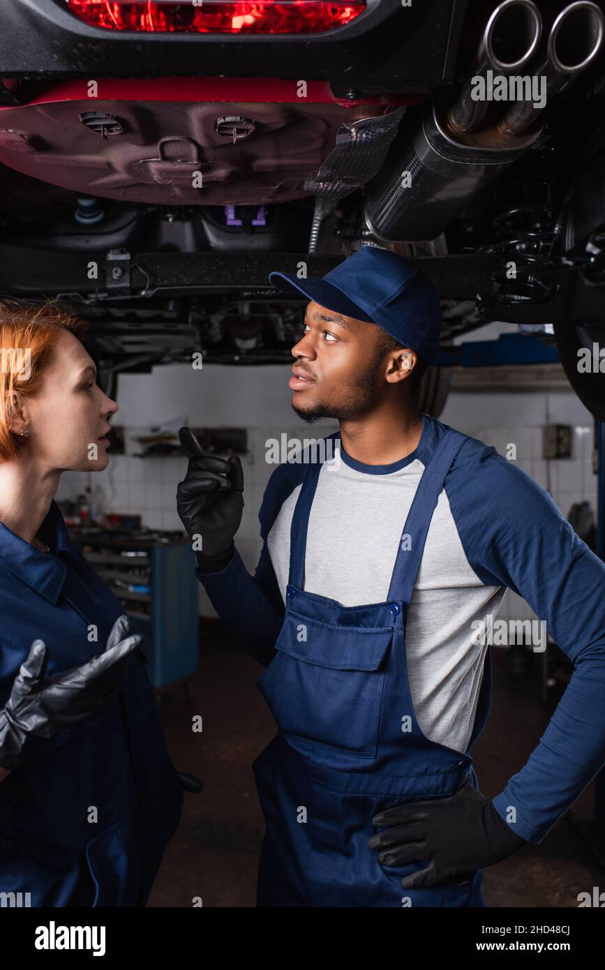 African american mechanic pointing at car bottom near colleague in ...