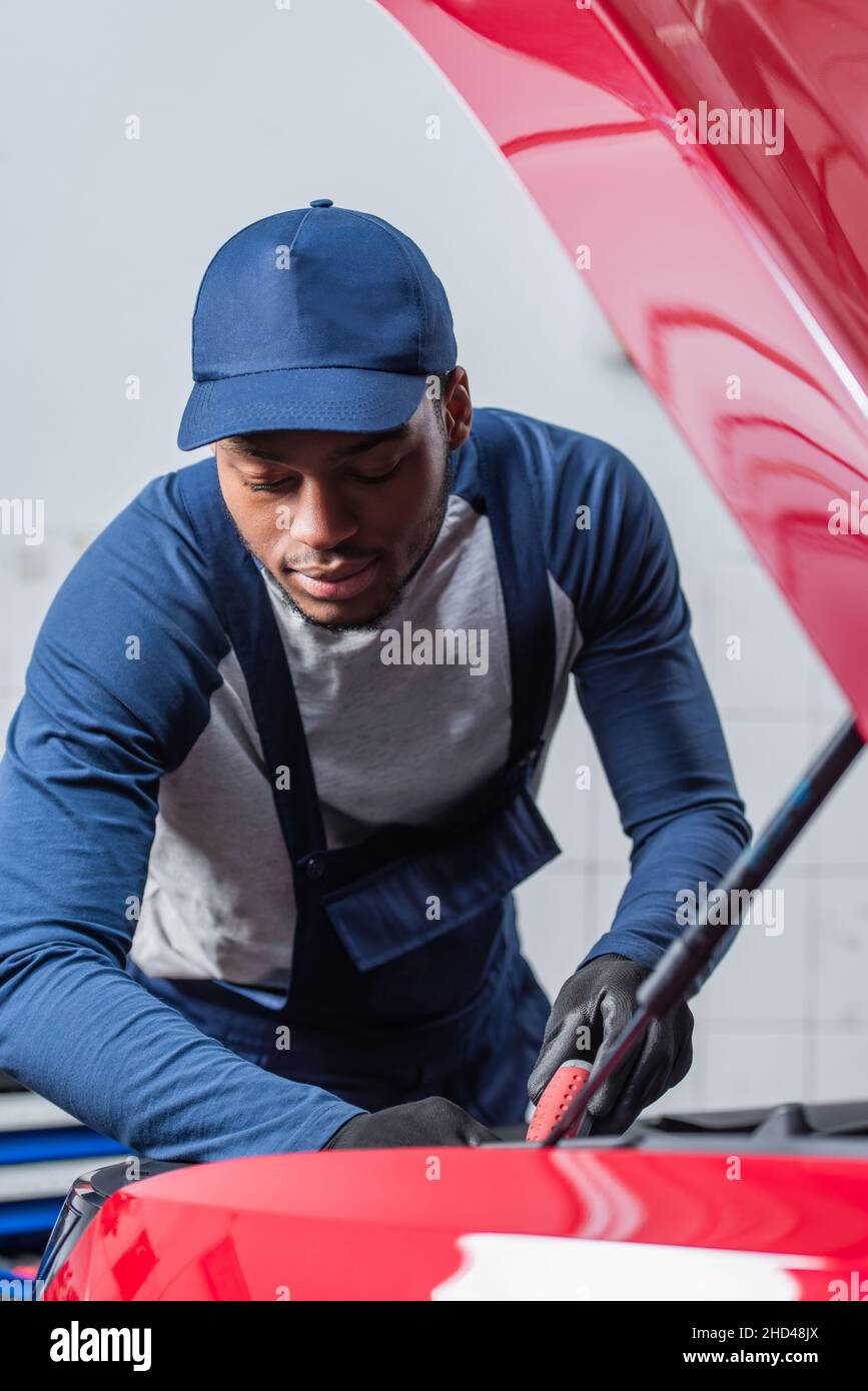 young african american technician in overalls and cap inspecting car ...