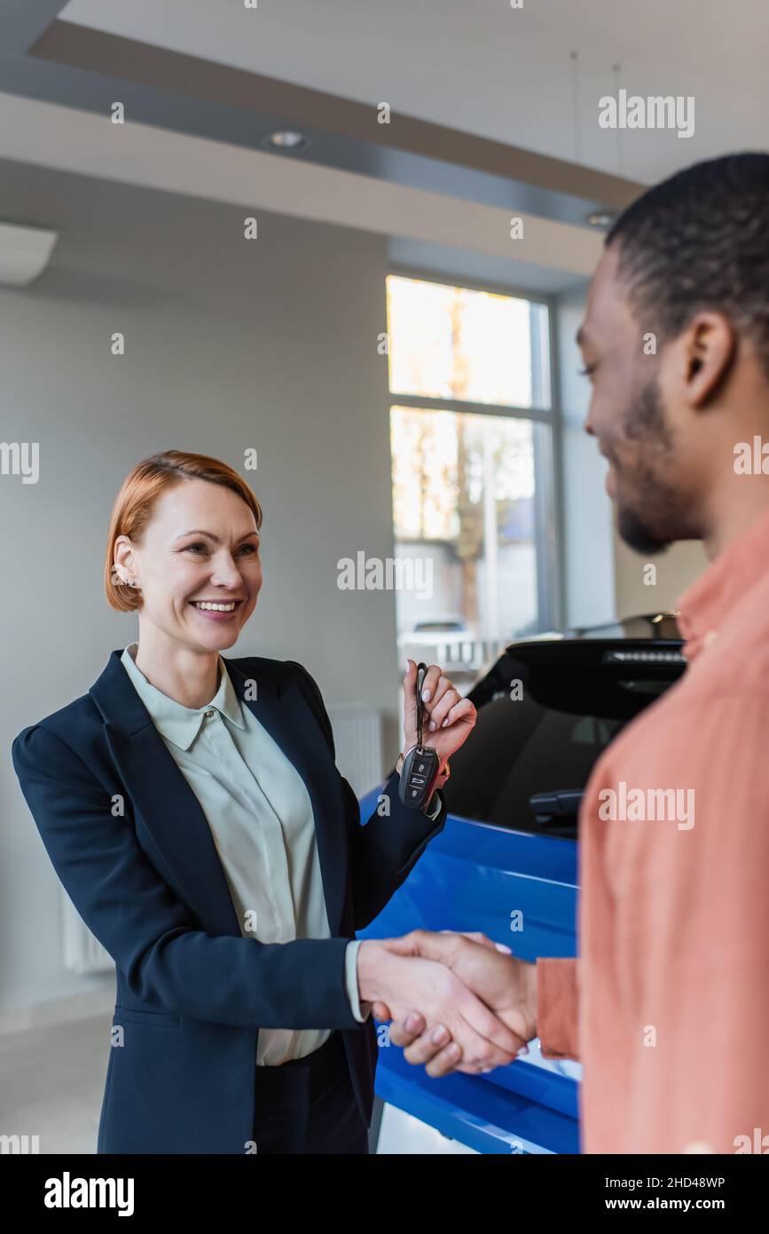 happy car dealer holding key and shaking hands with blurred african ...