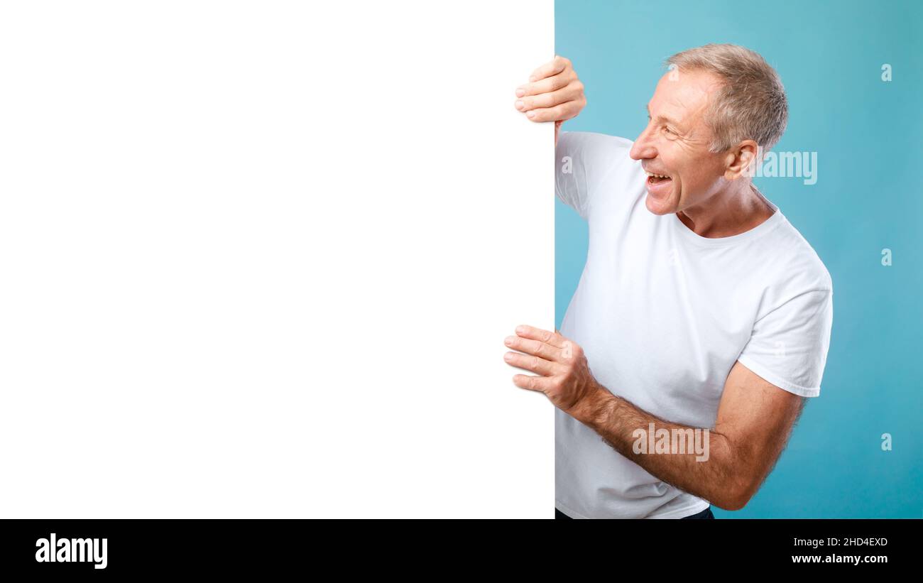 Mature man peeking out blank white advertising billboard at studio ...