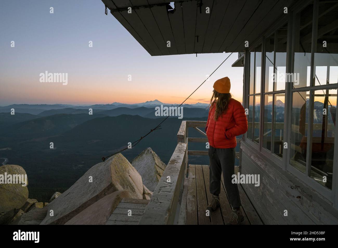 Female in a hat and puffy jacket standing on a fire lookout at sunset ...