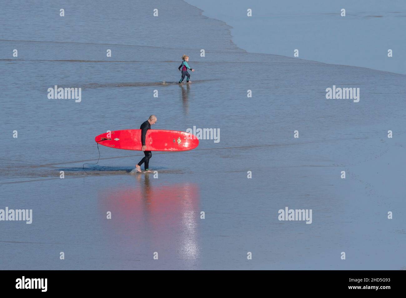 A child walking out of the sea and a tired surfer carrying a bright red ...