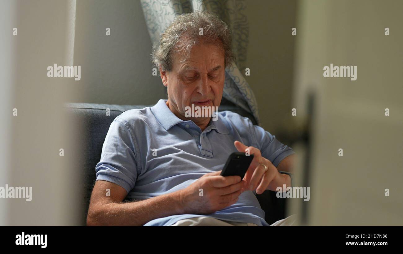 Candid old man using phone, senior person sitting on sofa looking at ...