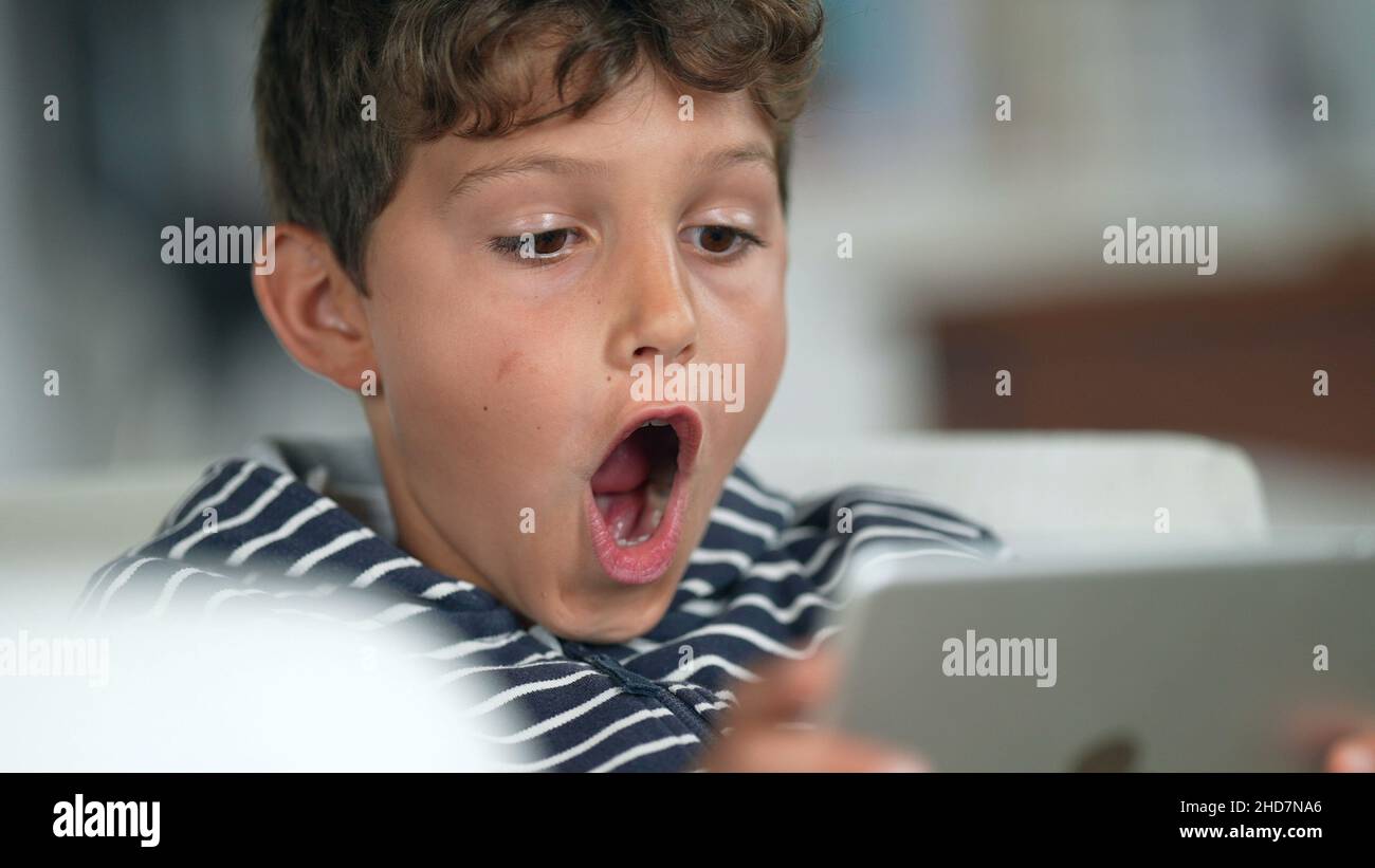Child excited reaction playing video-game. Boy shocking surprise open mouth reacting to screen ...