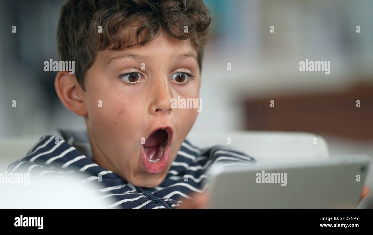 Child excited reaction playing video-game. Boy shocking surprise open mouth reacting to screen ...