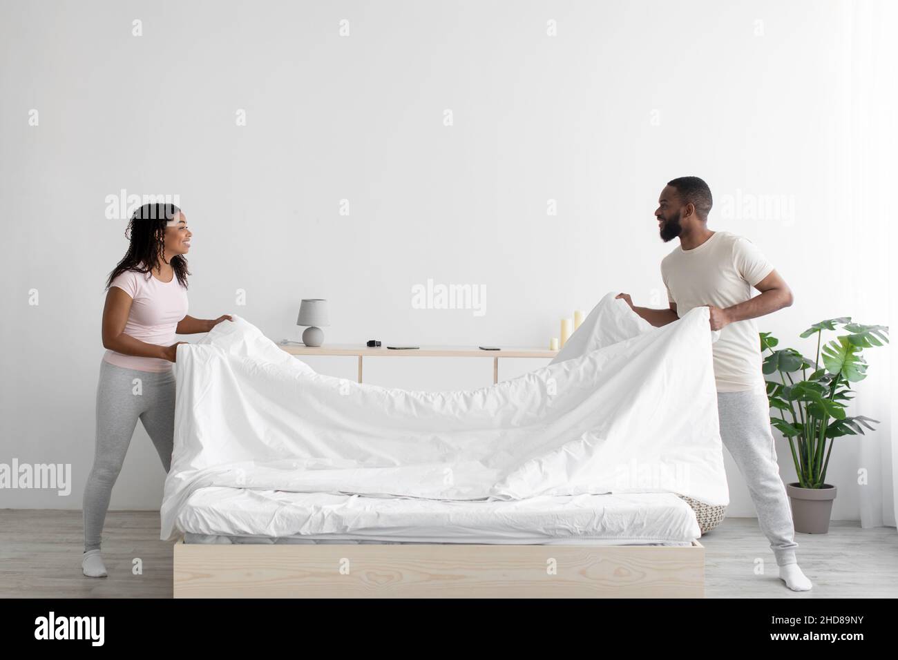 Cheerful young black woman and guy make bed and hold blanket in bedroom ...