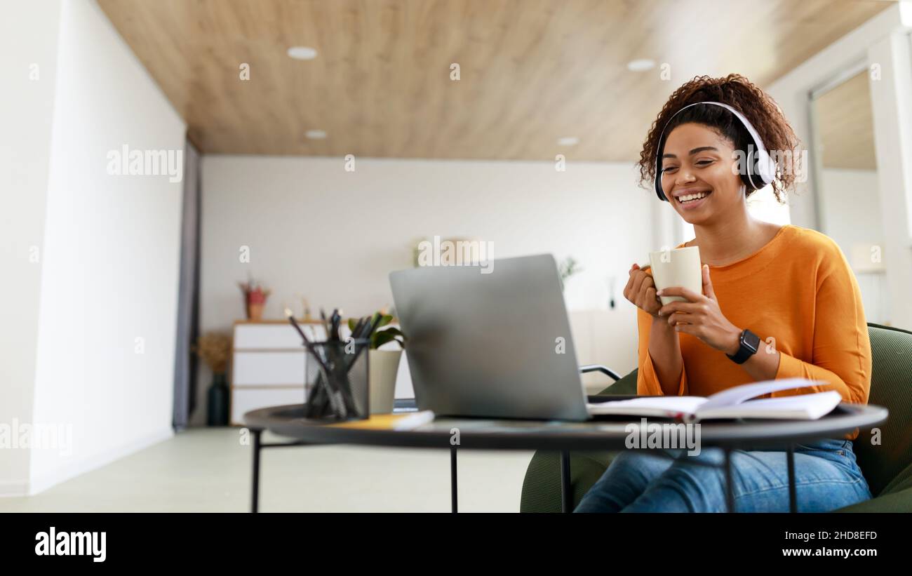 Smiling black lady watching video on computer, drinking hot coffee ...