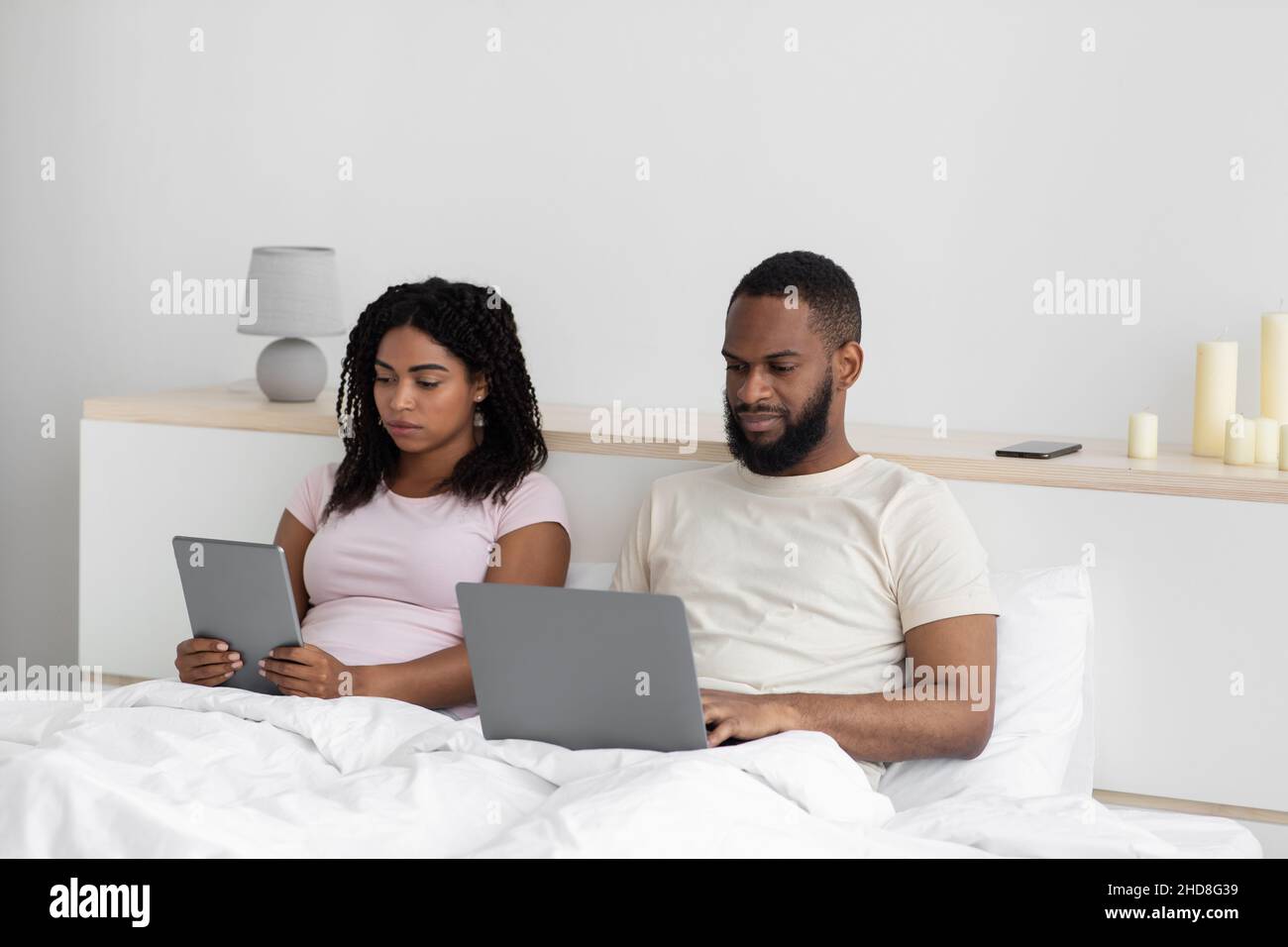 Busy serious millennial black wife and husband working on laptop and tablet, sitting on bed ...