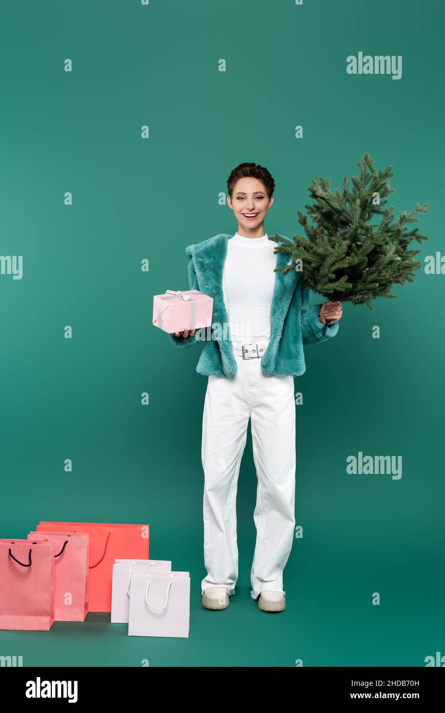 full length view of pleased woman with gift box and small pine tree ...