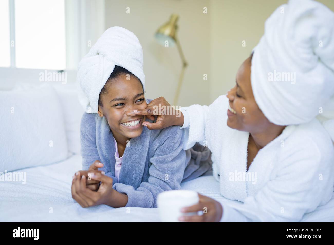 Happy Mother Applying Skin Cream On Nose Of Daughter In Bathrobe On Bed