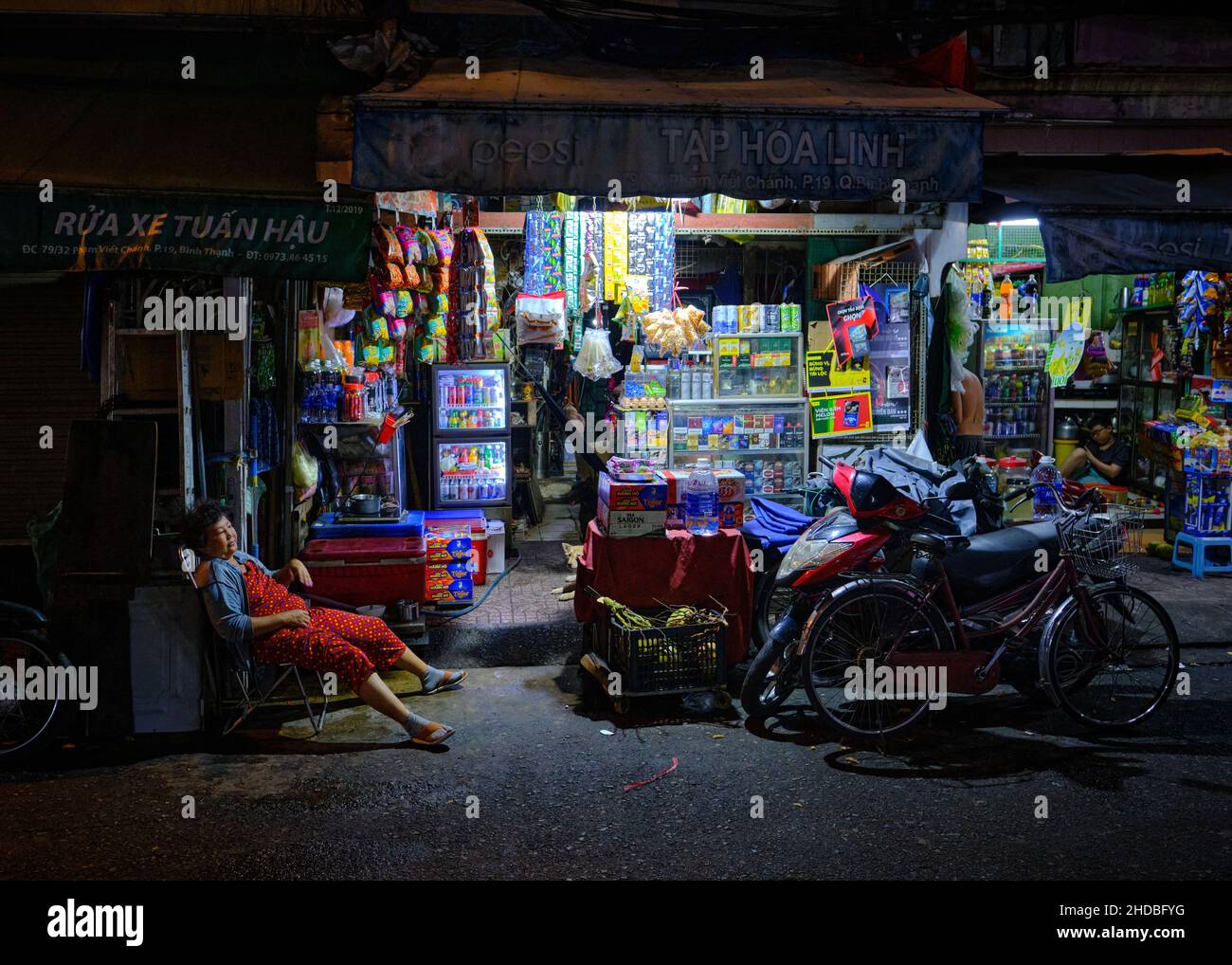 Saigon corner store at night—illuminated by light, with a relaxed lady ...
