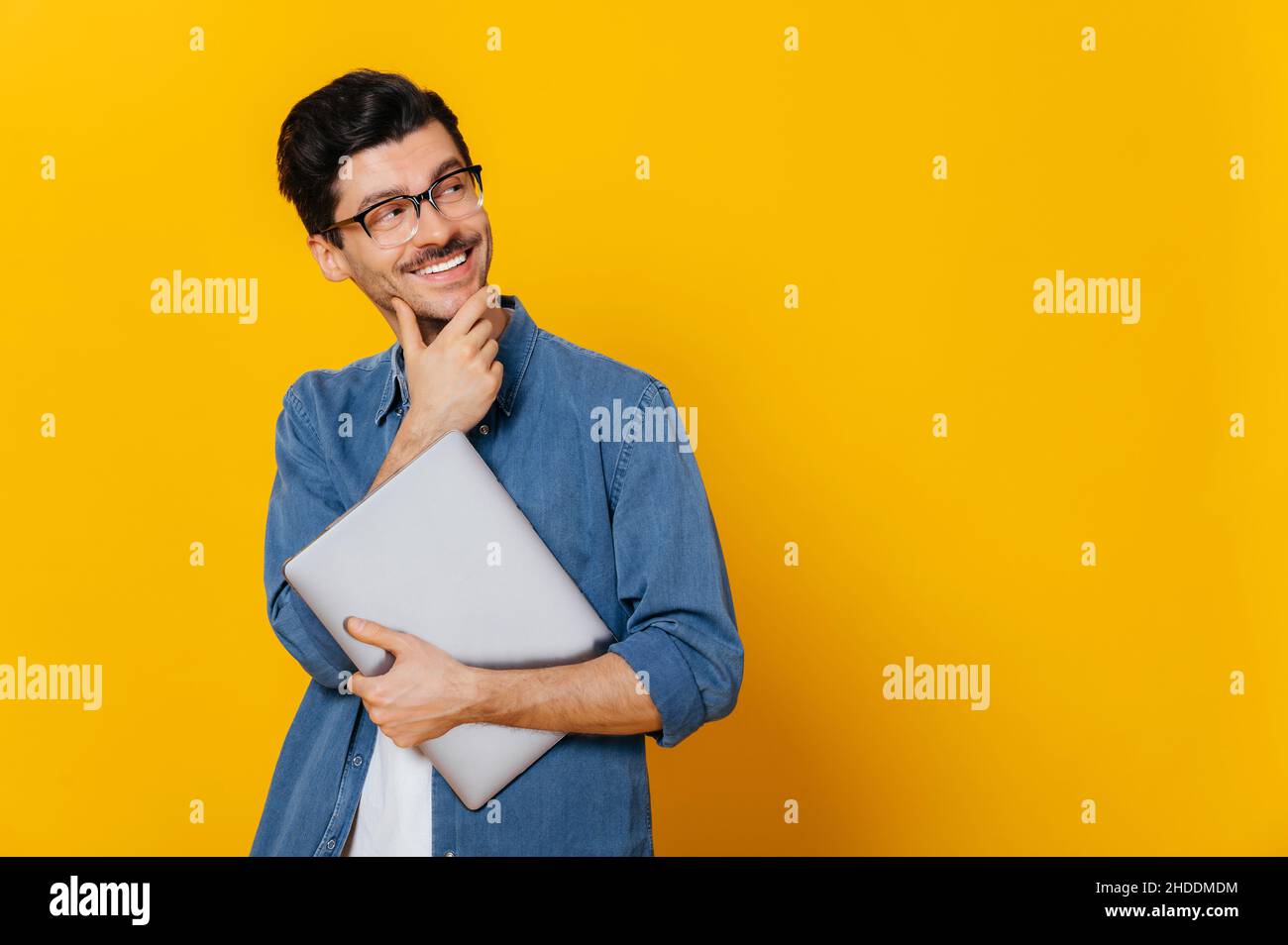 Positive smart guy in glasses and in a denim shirt, holding a laptop at ...