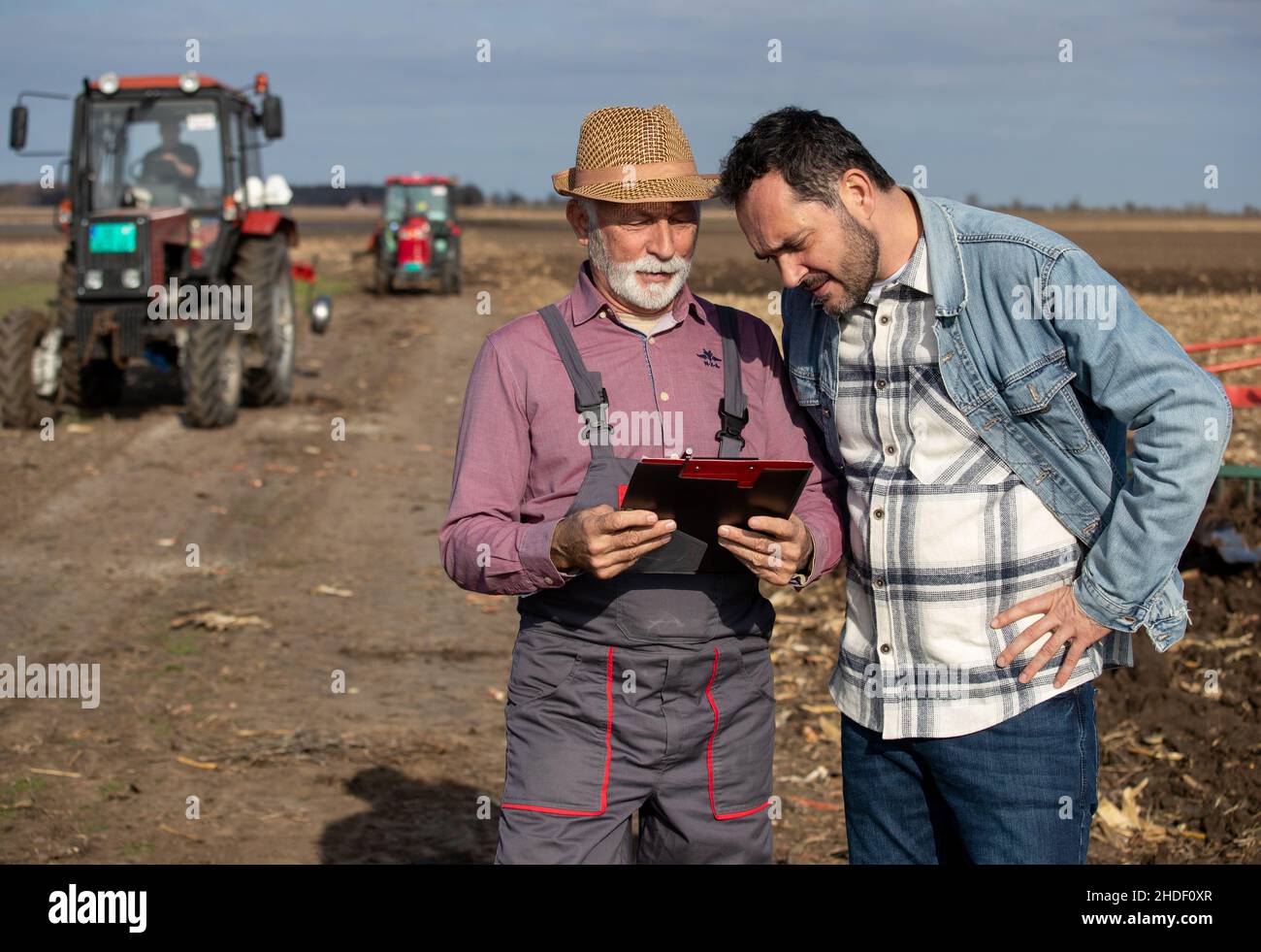 Two farmers standing in field and looking at notepad. Tractors working ...