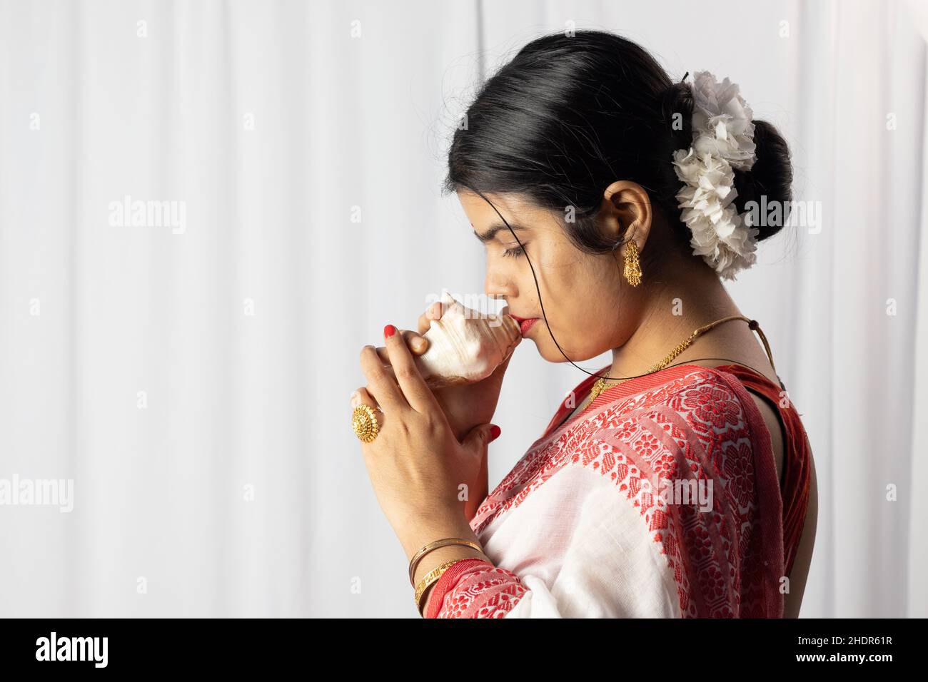 A beautiful Indian woman in red saree blowing conch shell on white ...