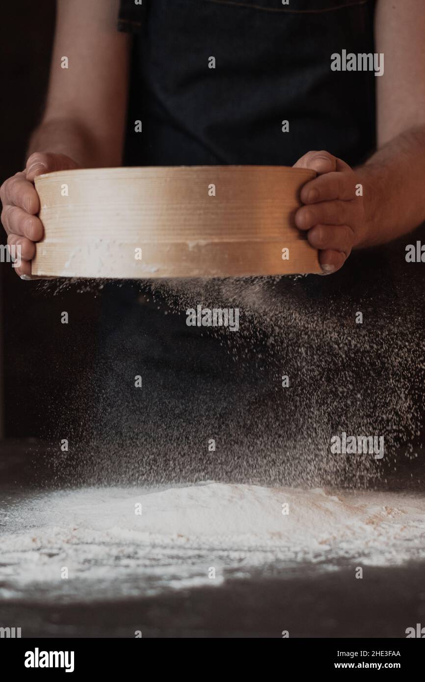 A dark background baker's hands sift white wheat flour over a sieve ...