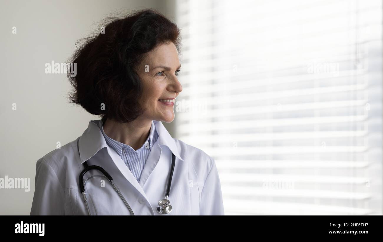 Dreamy smiling beautiful female GP doctor looking out of window Stock ...