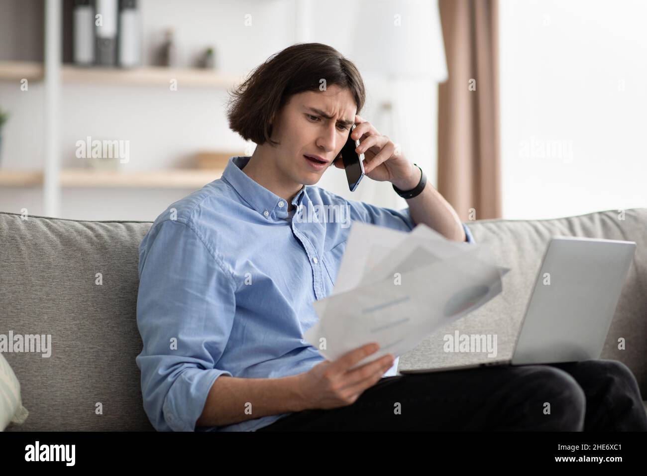 Angry young man working from home, sitting on sofa with laptop, holding ...