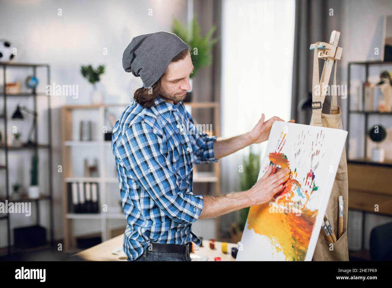 Skilful male artist drawing with colorful paintings on easel at home ...