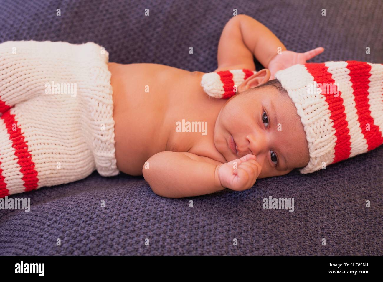 Cute lying newborn baby in a striped hat looks looks up with his hand ...