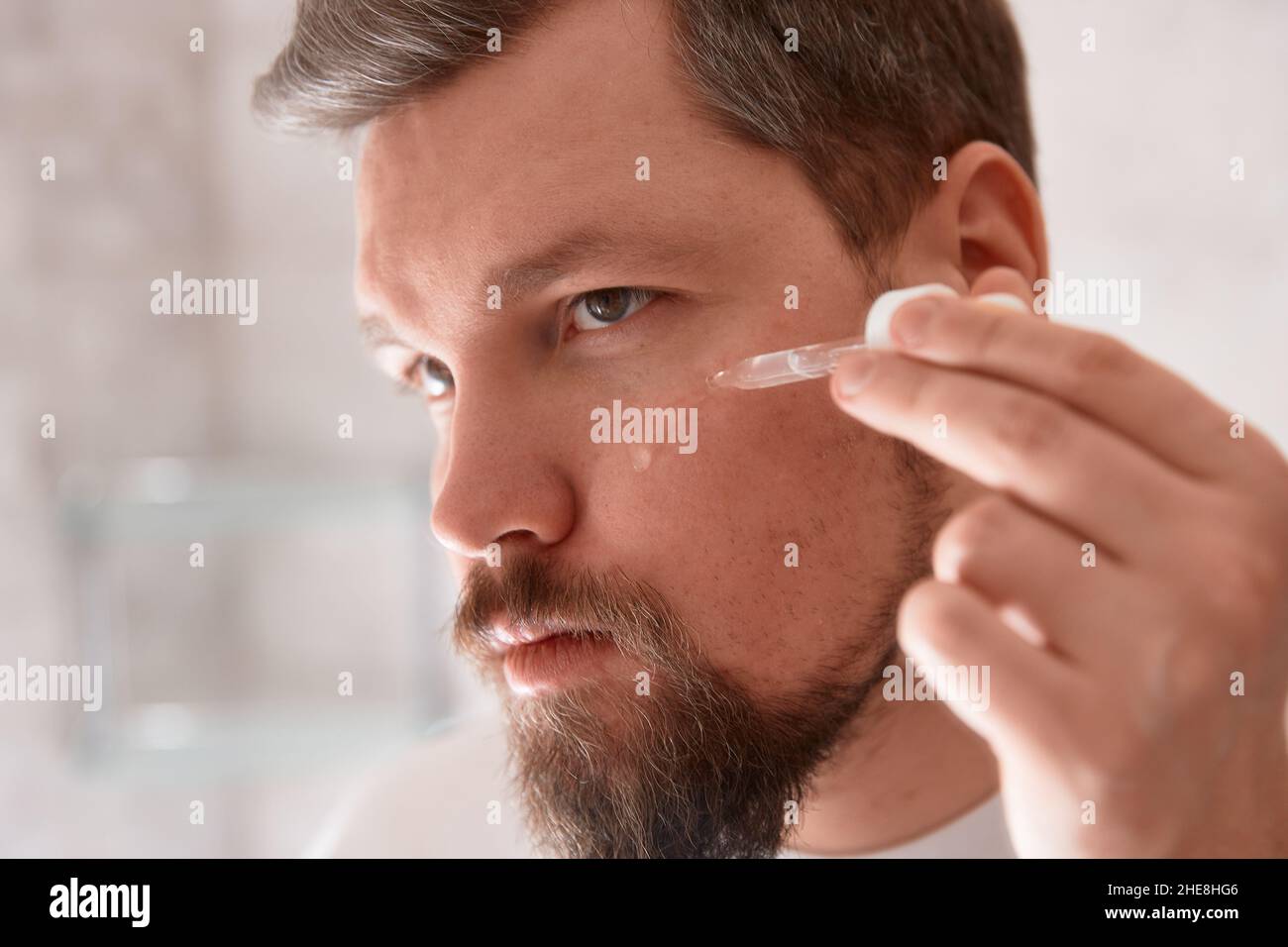 Portrait of man in white tshirt applying serum on his face at bathroom ...