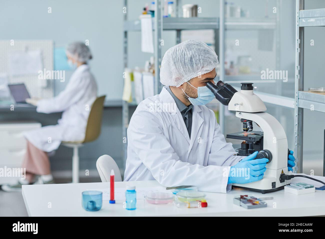 Young scientist in lab coat and protective mask examining sample of new ...