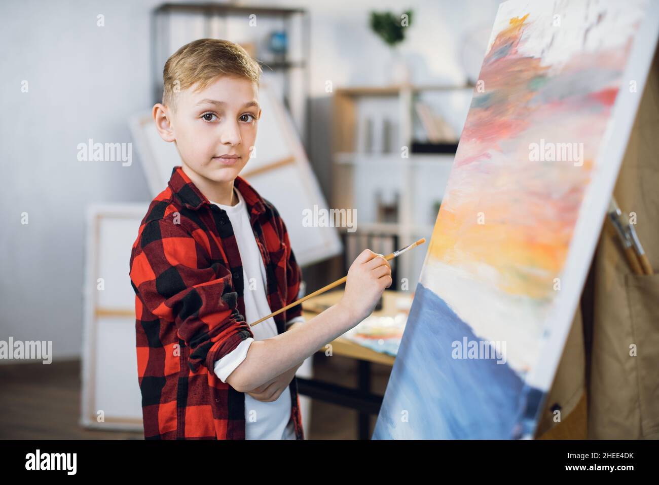 Portrait of talented cute boy drawing with brush and paints on easel ...