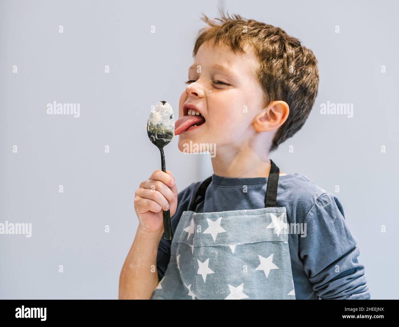 Funny little kid with brown hair and in apron licking spoon while ...