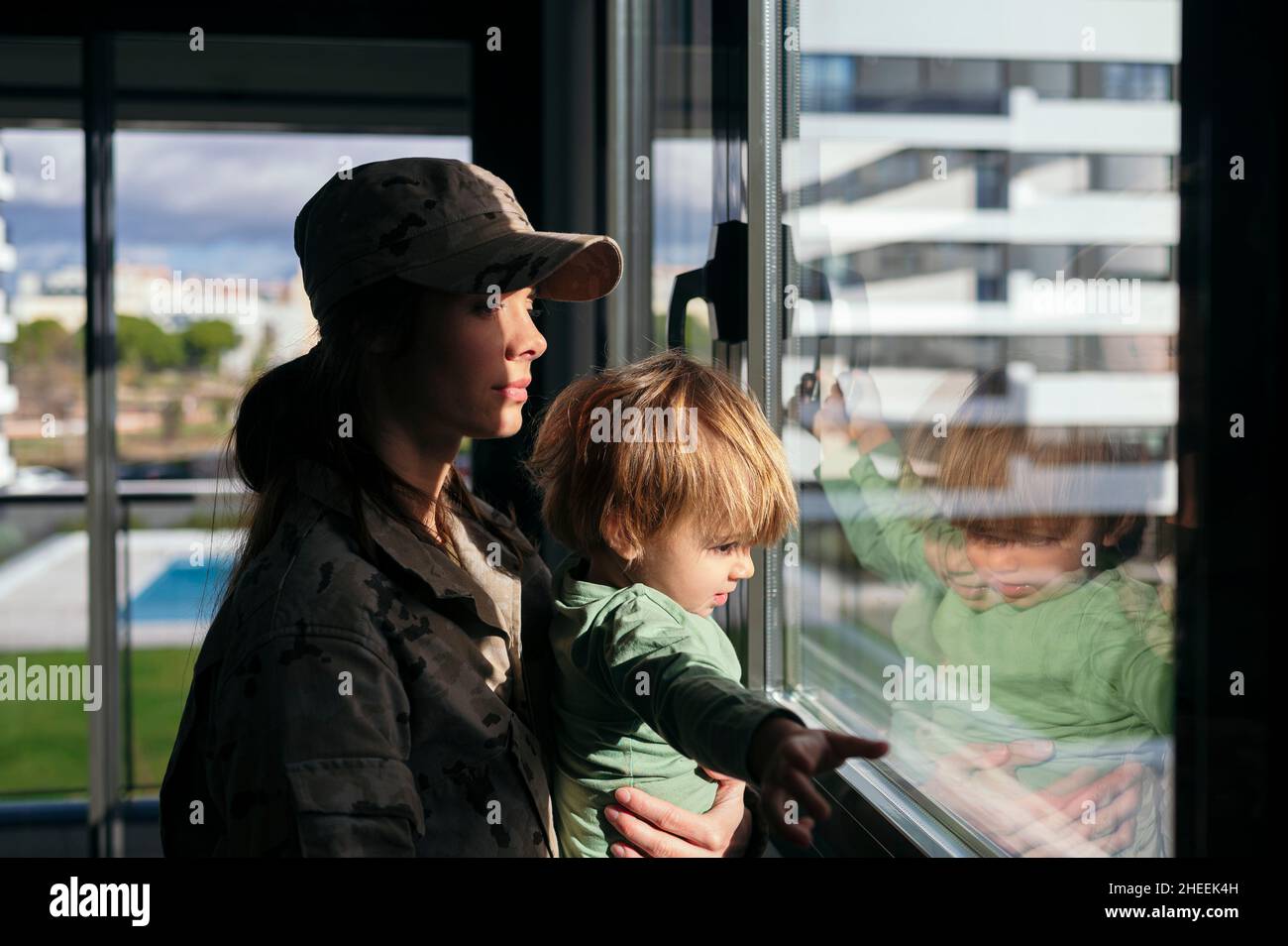Side view of female soldier in uniform looking out window with cute son ...