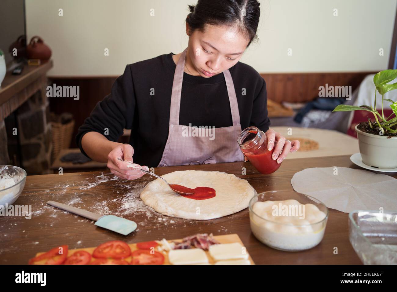 Asian cook in apron pouring tomato sauce on dough while making homemade ...