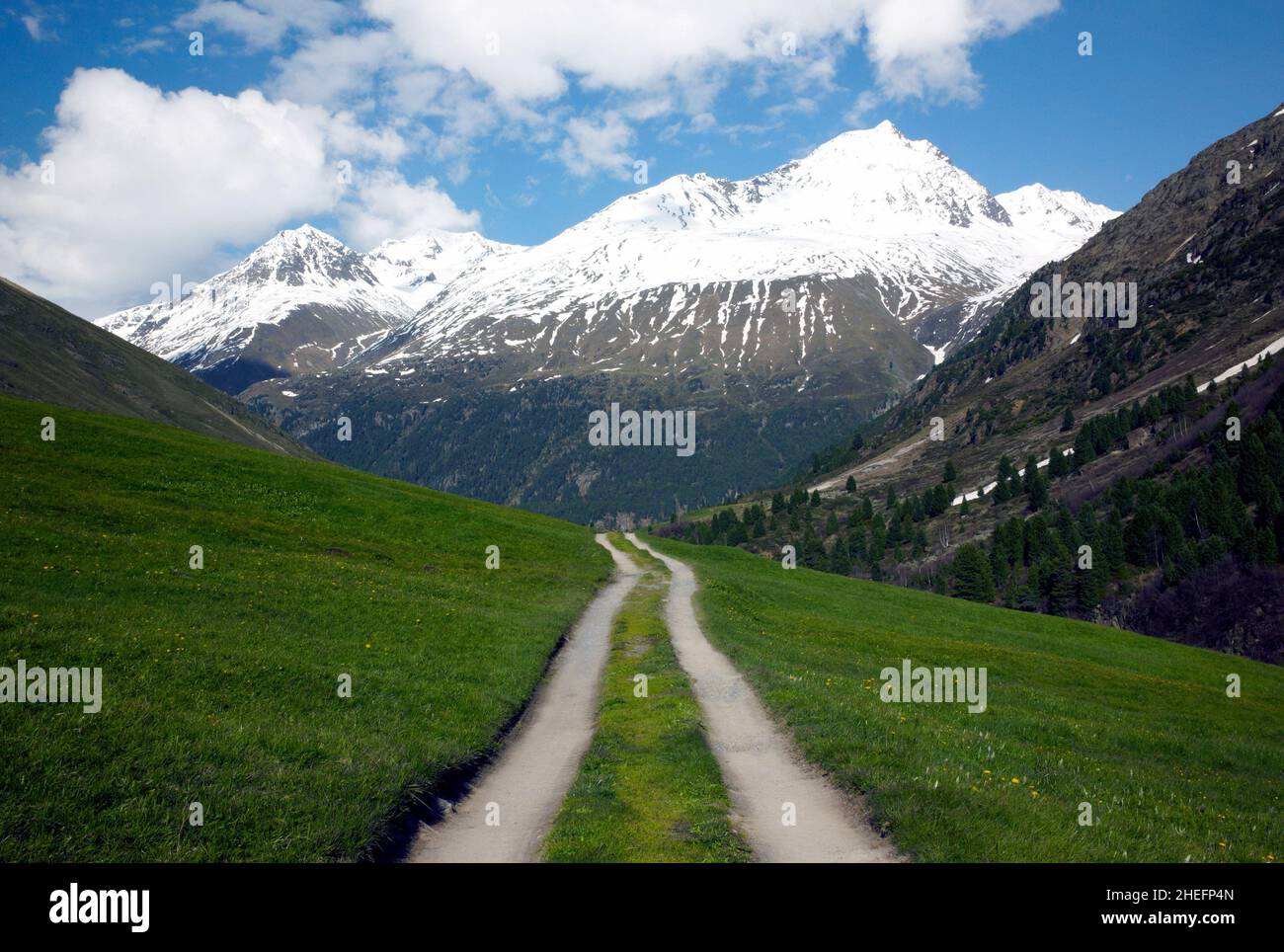 Colour photograph of a mountain valley track, green fields and snow ...