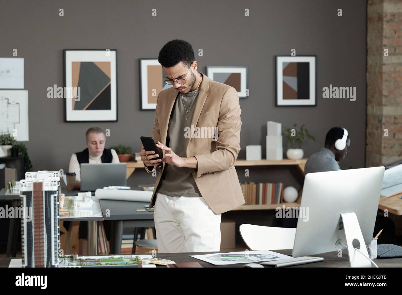 Young biracial businessman scrolling in smartphone while standing by workplace with computer ...