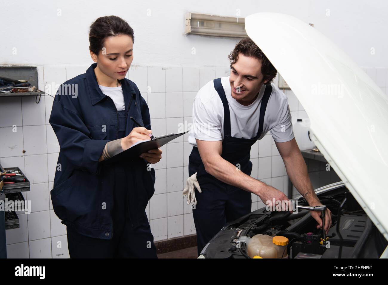 Mechanic writing on clipboard near smiling colleague and car in garage ...