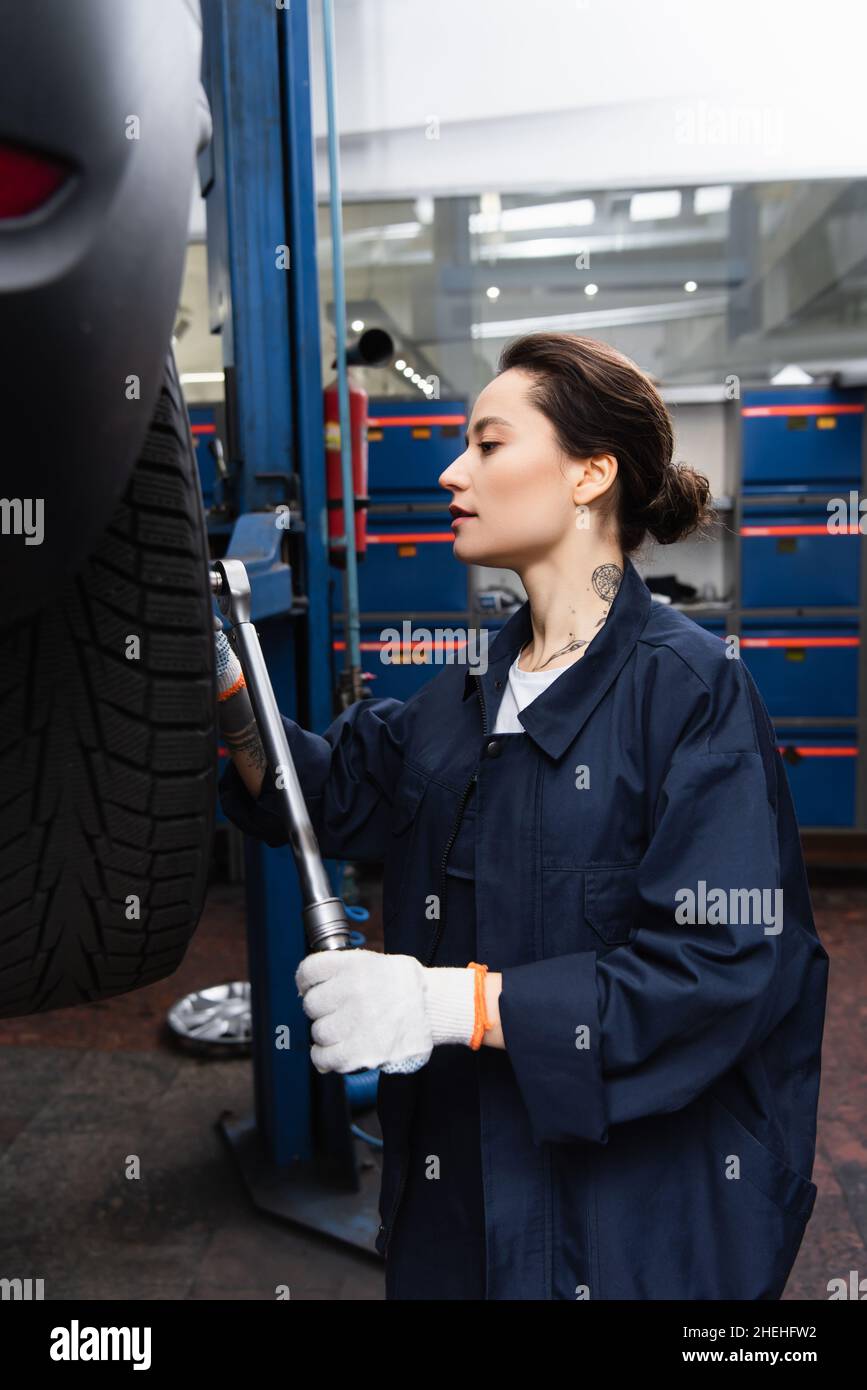 Side view of forewoman in uniform holding wrench near wheel of auto in ...