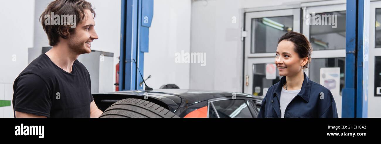 Smiling mechanic looking at colleague near tire and car in service ...