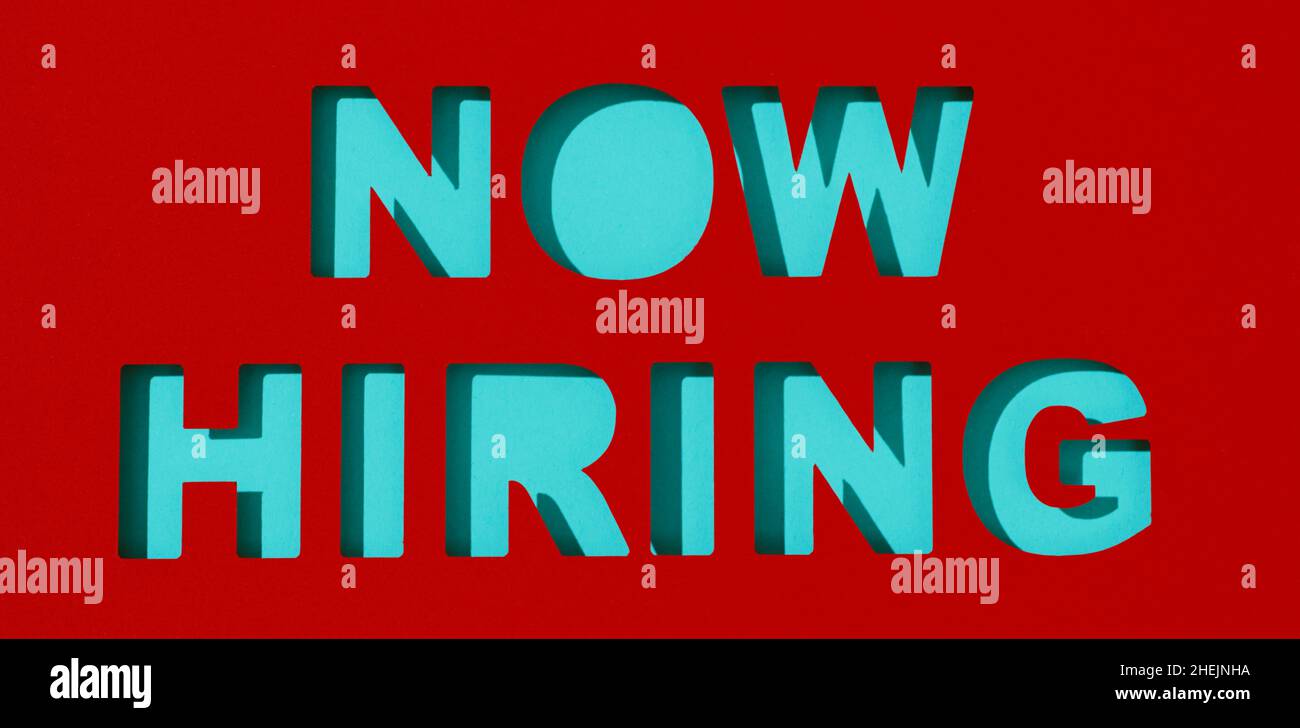 text now hiring cut out on a red paper, over a blue background, in a ...