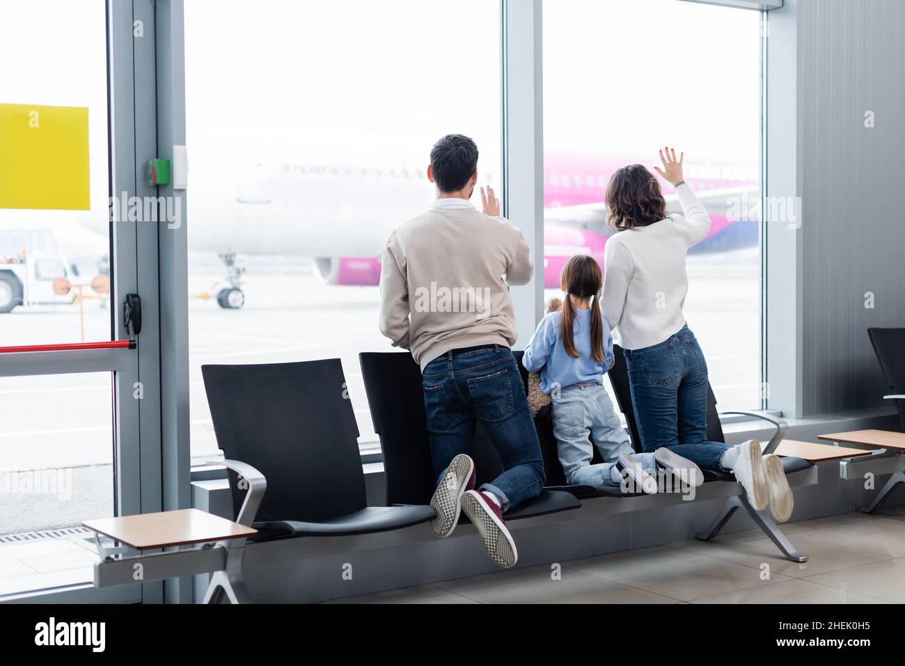 back view of family waving hands while looking at plane through window ...