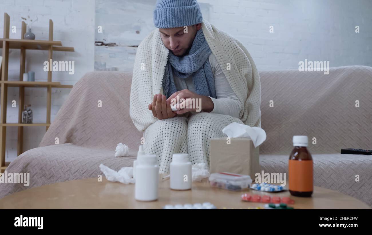 sick man in warm blanket and hat holding pills container near table ...