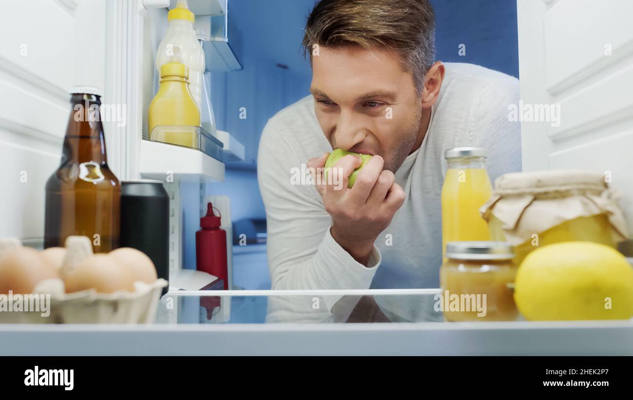 man eating ripe apple near fridge with eggs, beverages, containers and ...