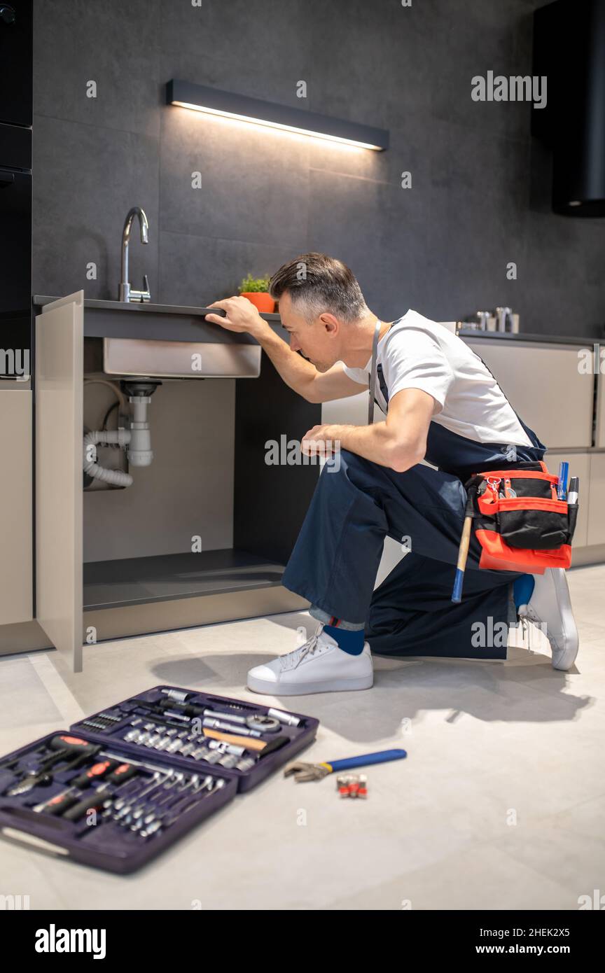 Man kneeling down touching sink inspecting pipes Stock Photo - Alamy
