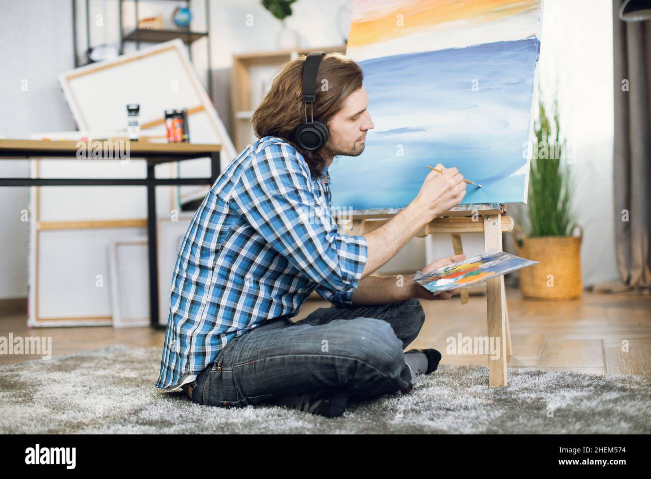 Happy caucasian man sitting on floor in wireless headset and painting ...