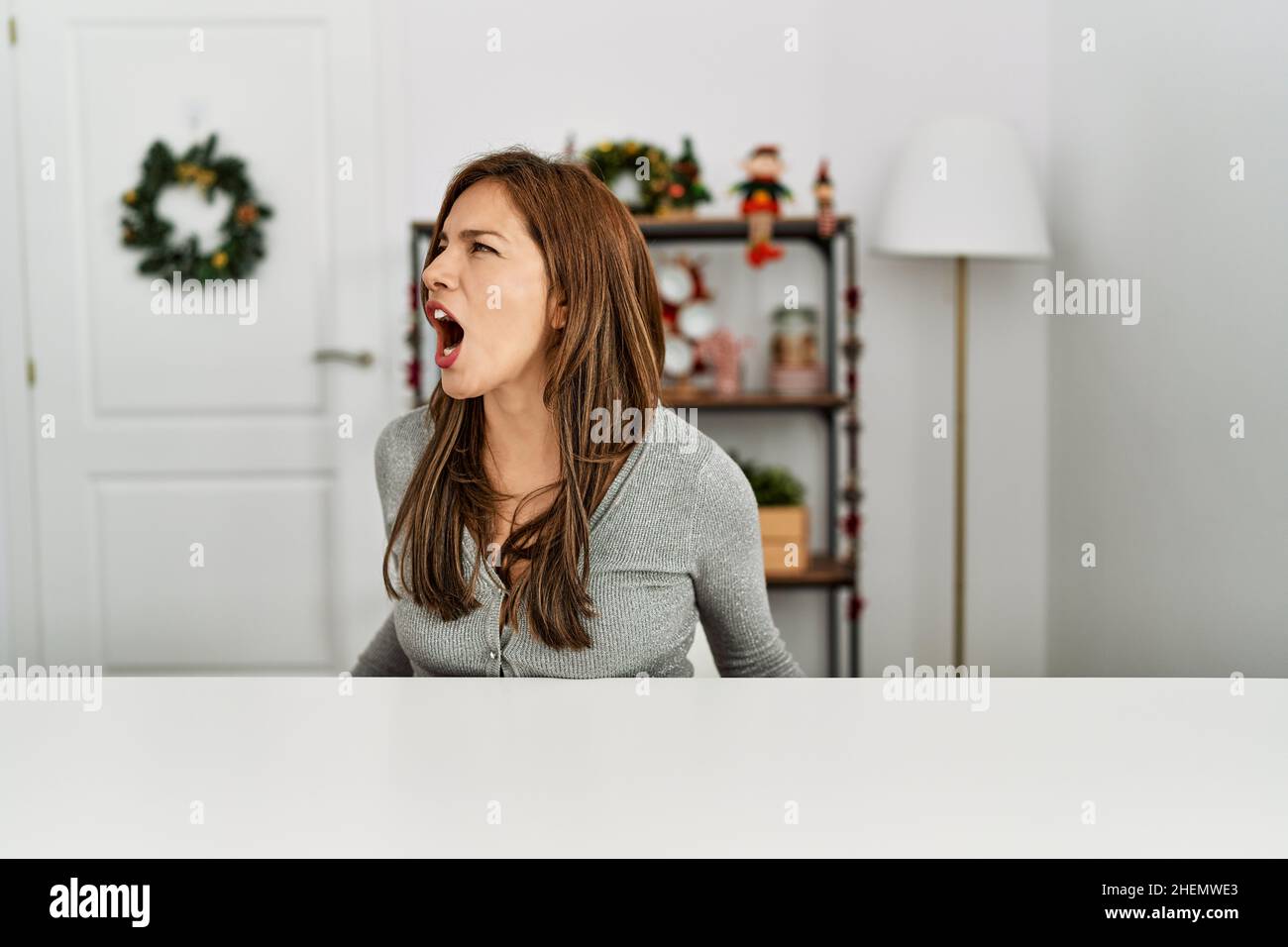 Young latin woman sitting on the table by christmas decor angry and mad ...