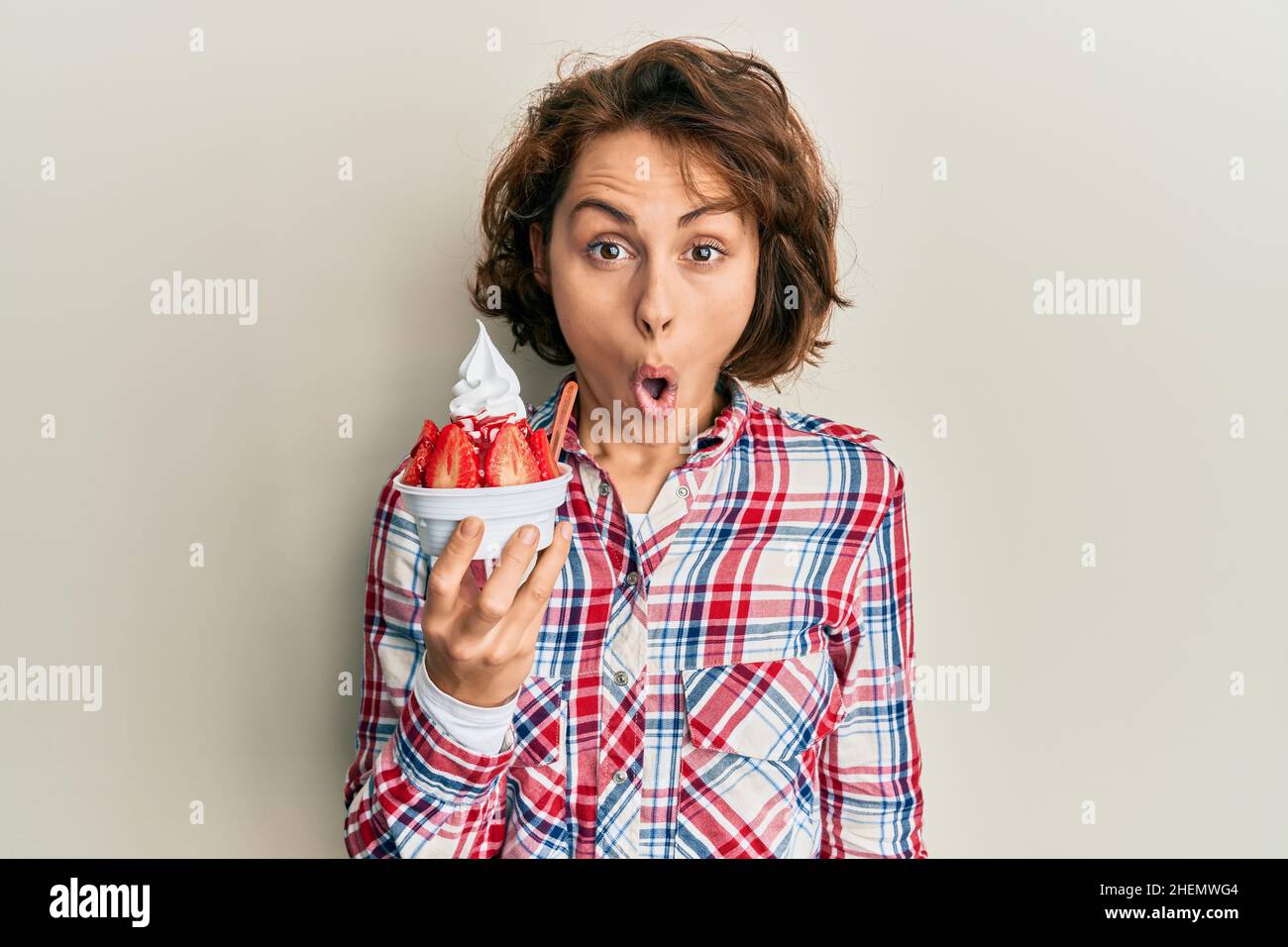 Young brunette woman eating strawberry ice cream scared and amazed with ...