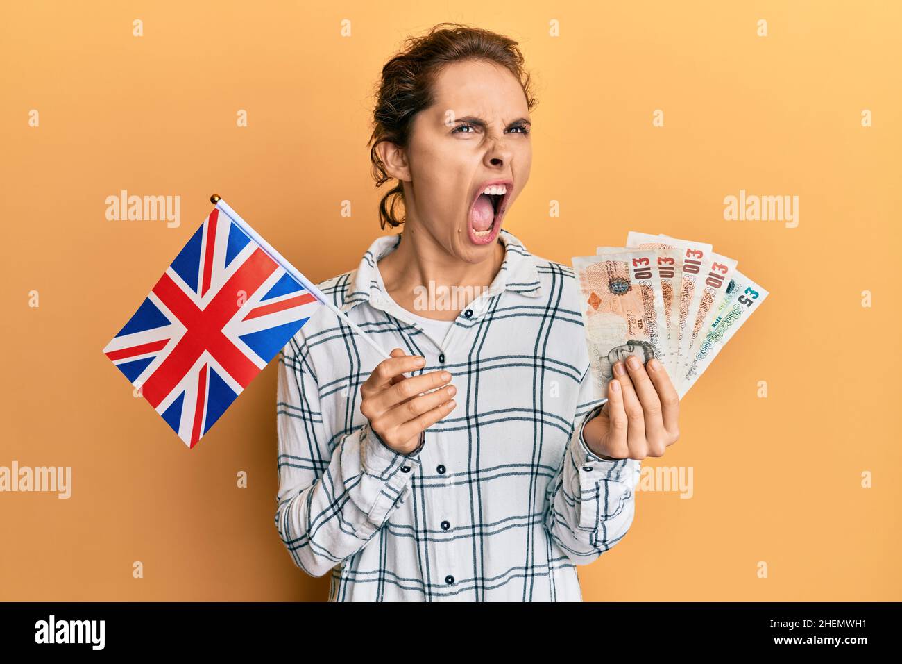 Young brunette woman holding uk flag and pounds banknotes angry and mad ...