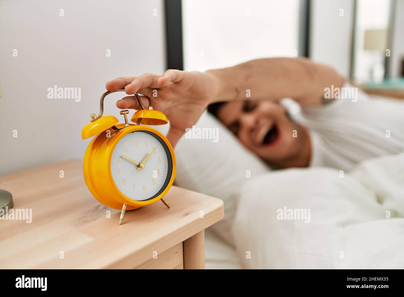 Young Hispanic Man Turning Off Alarm Clock Lying On The Bed At Bedroom