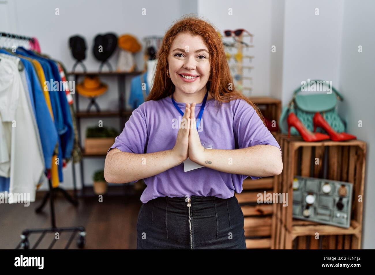 Young redhead woman working as manager at retail boutique praying with hands together asking for ...