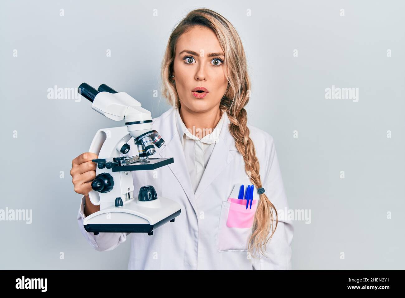 Beautiful young blonde woman holding microscope scared and amazed with ...