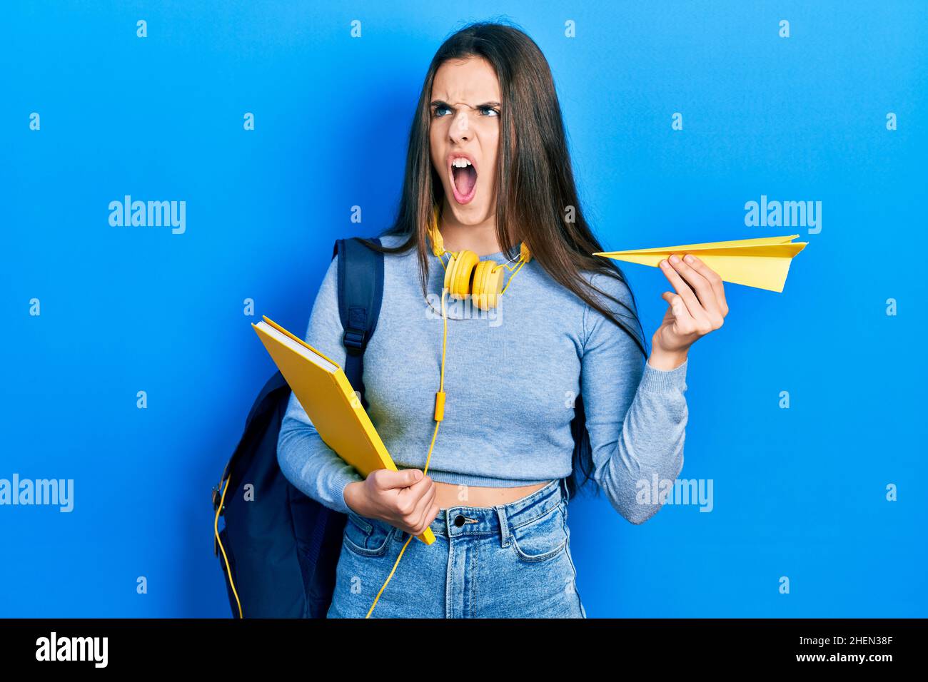 Young brunette teenager student holding books and paper plane angry and ...