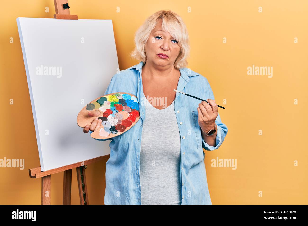 Middle age blonde woman standing drawing with palette by painter easel ...